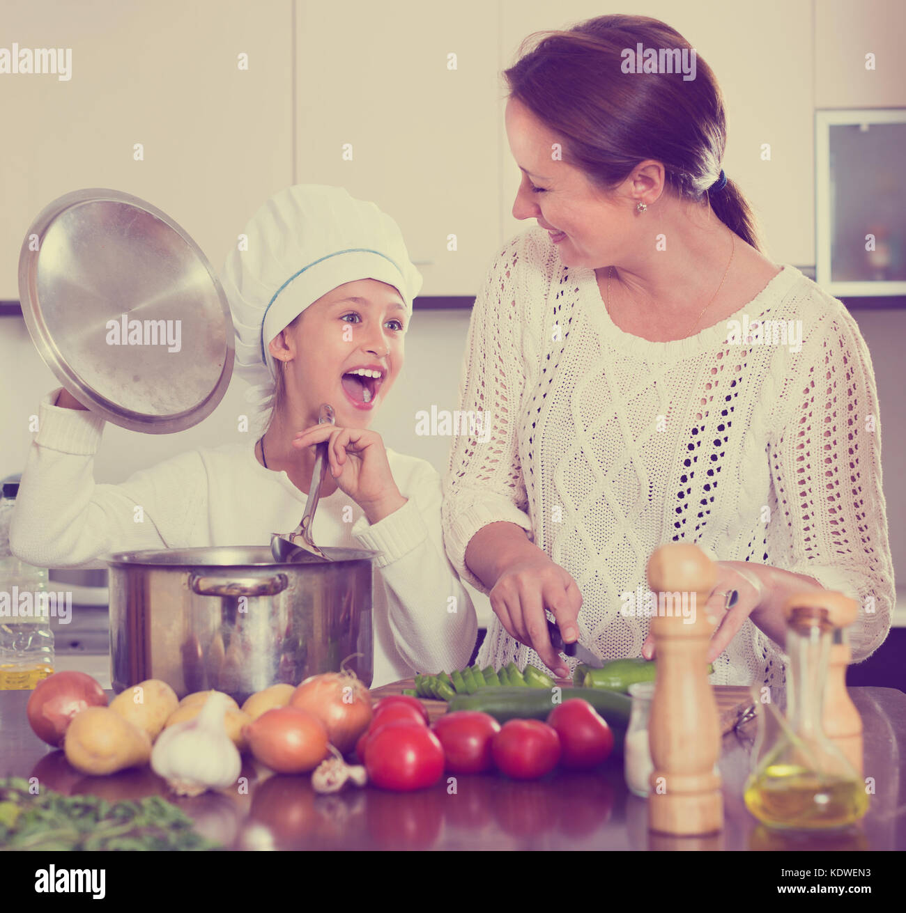 Mother and her cheerful daughter in cookers’s hat preparing soup ...