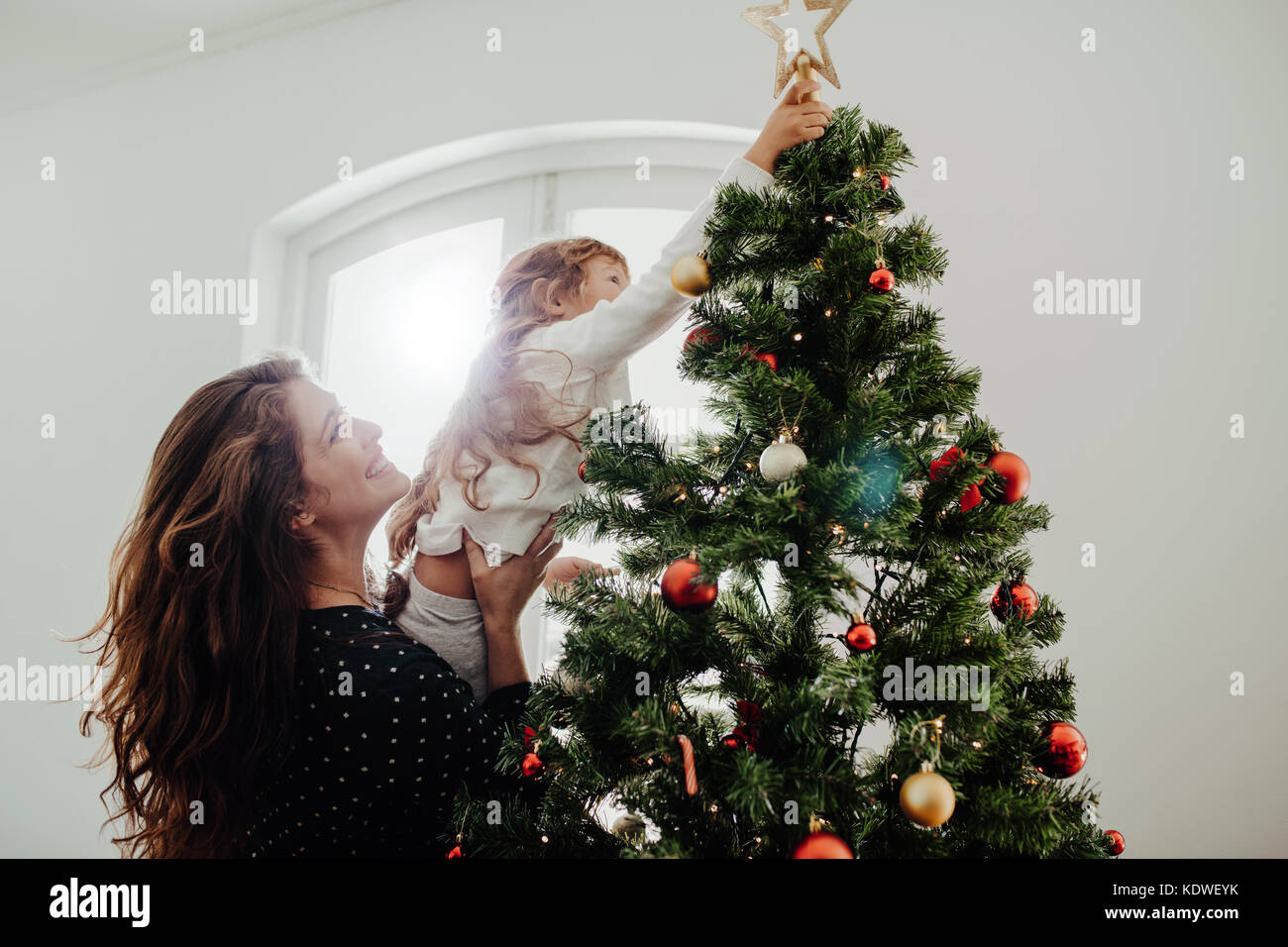 Little girl placing a star on the top of Christmas tree. Mother lifting ...