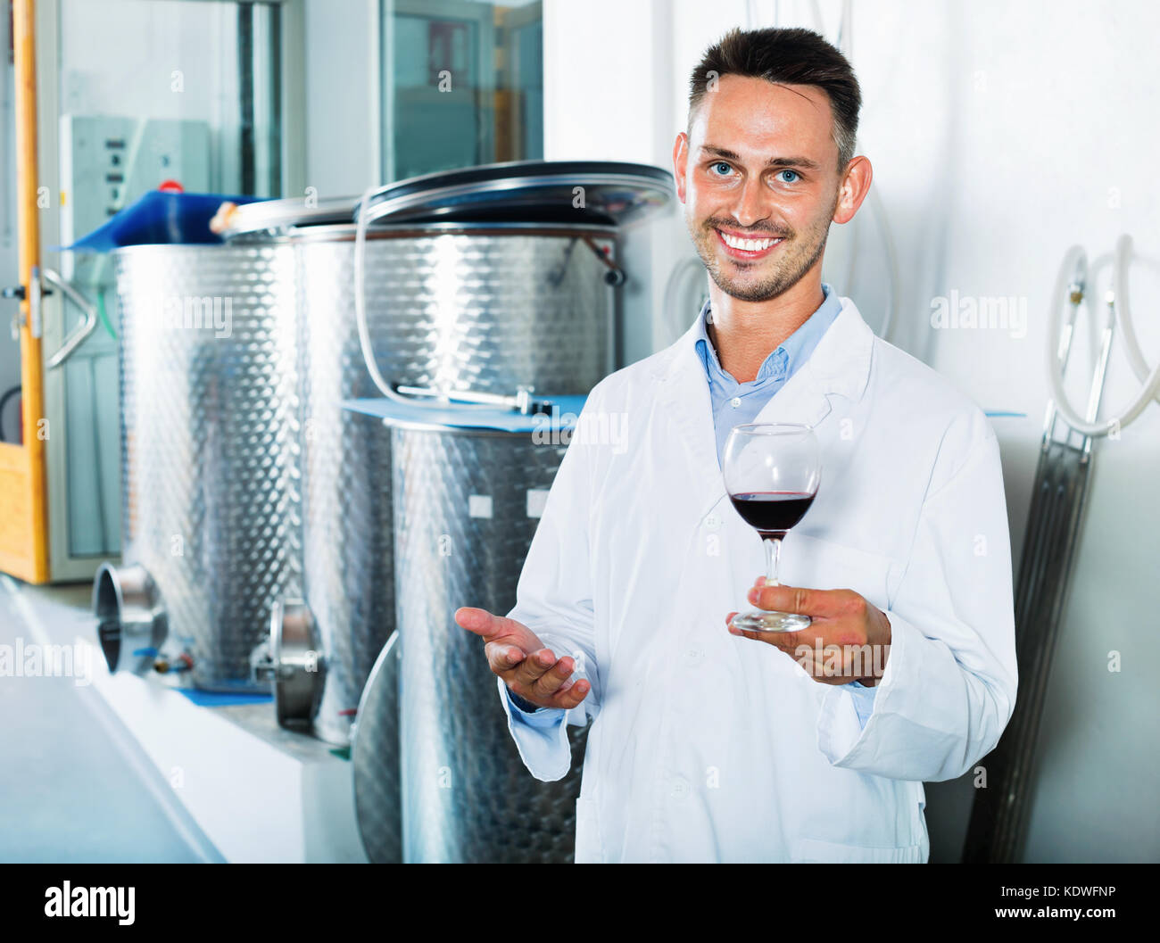 Smiling male young winemaker in lab coat having glass of wine in hands ...