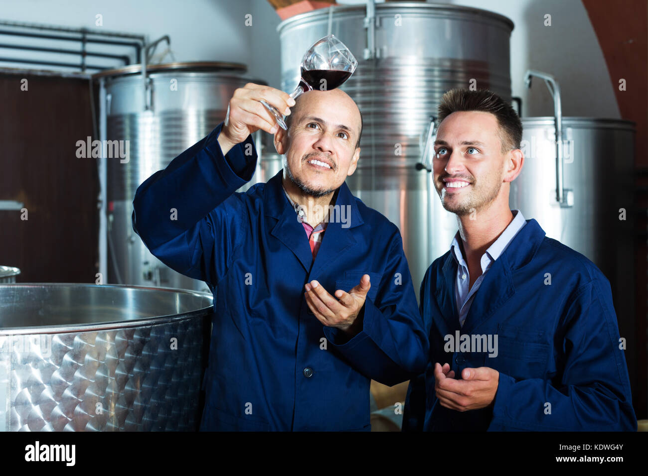 two cheerful winery male workers in uniform holding wine sample in ...