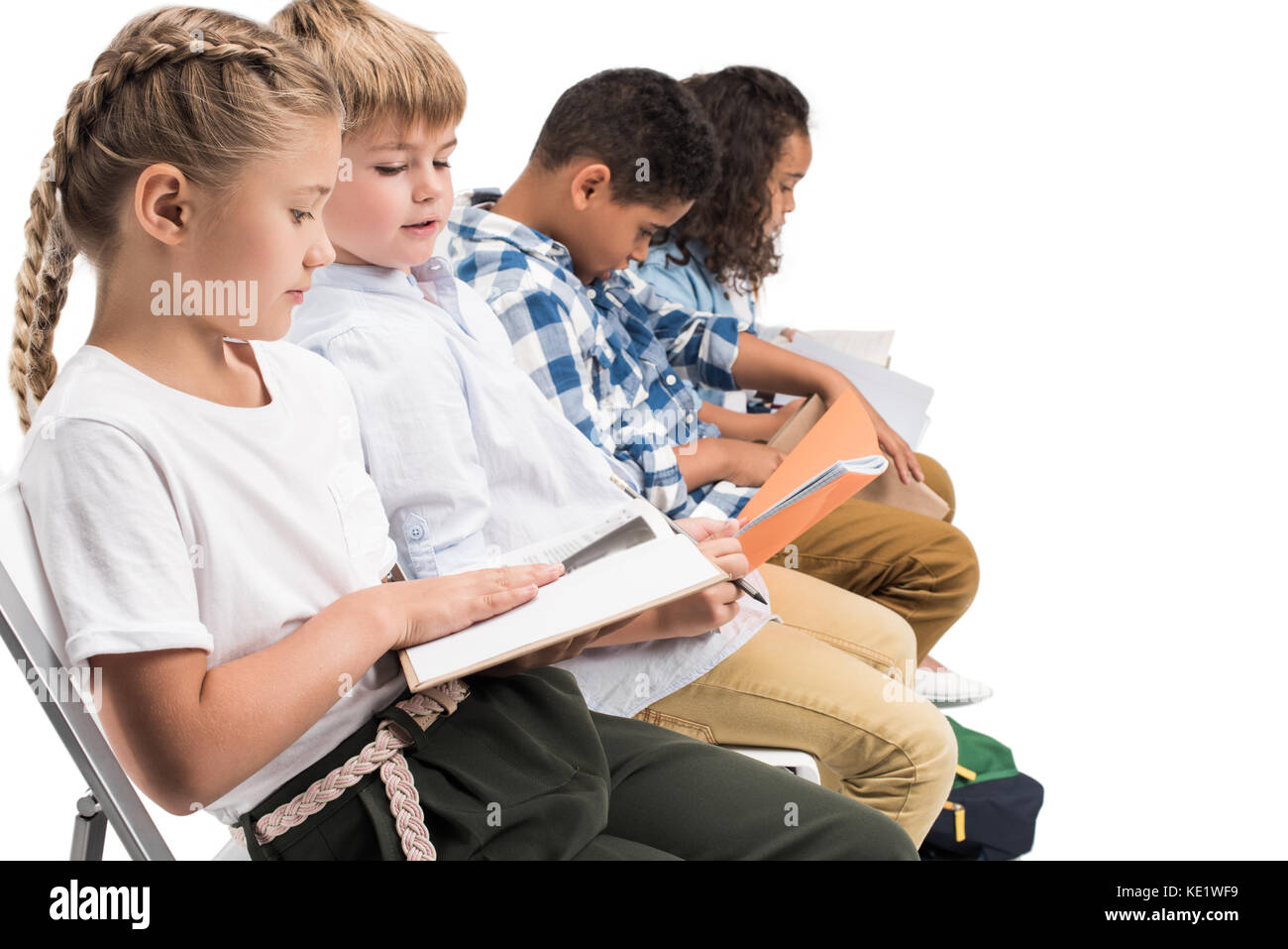 multiethnic kids reading books while sitting on chairs isolated on ...