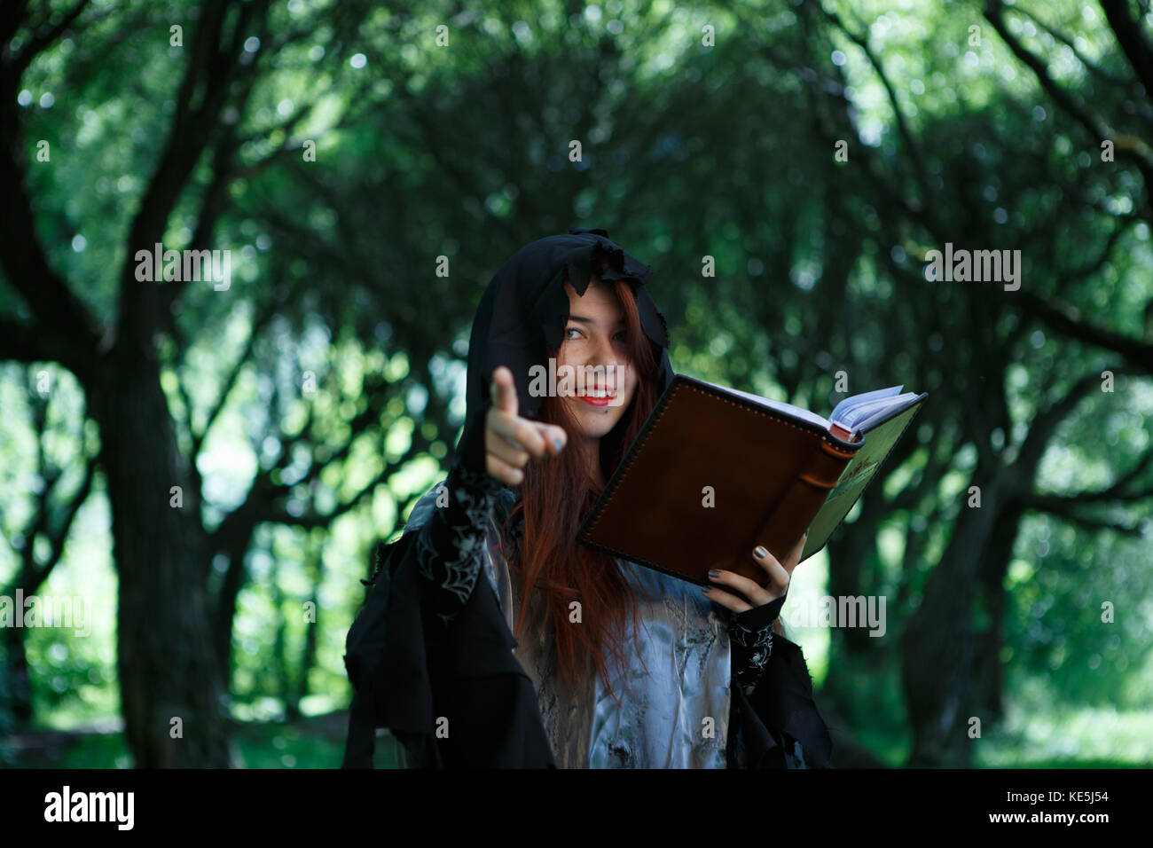 Portrait of smiling witch with book pointing finger at camera in ...
