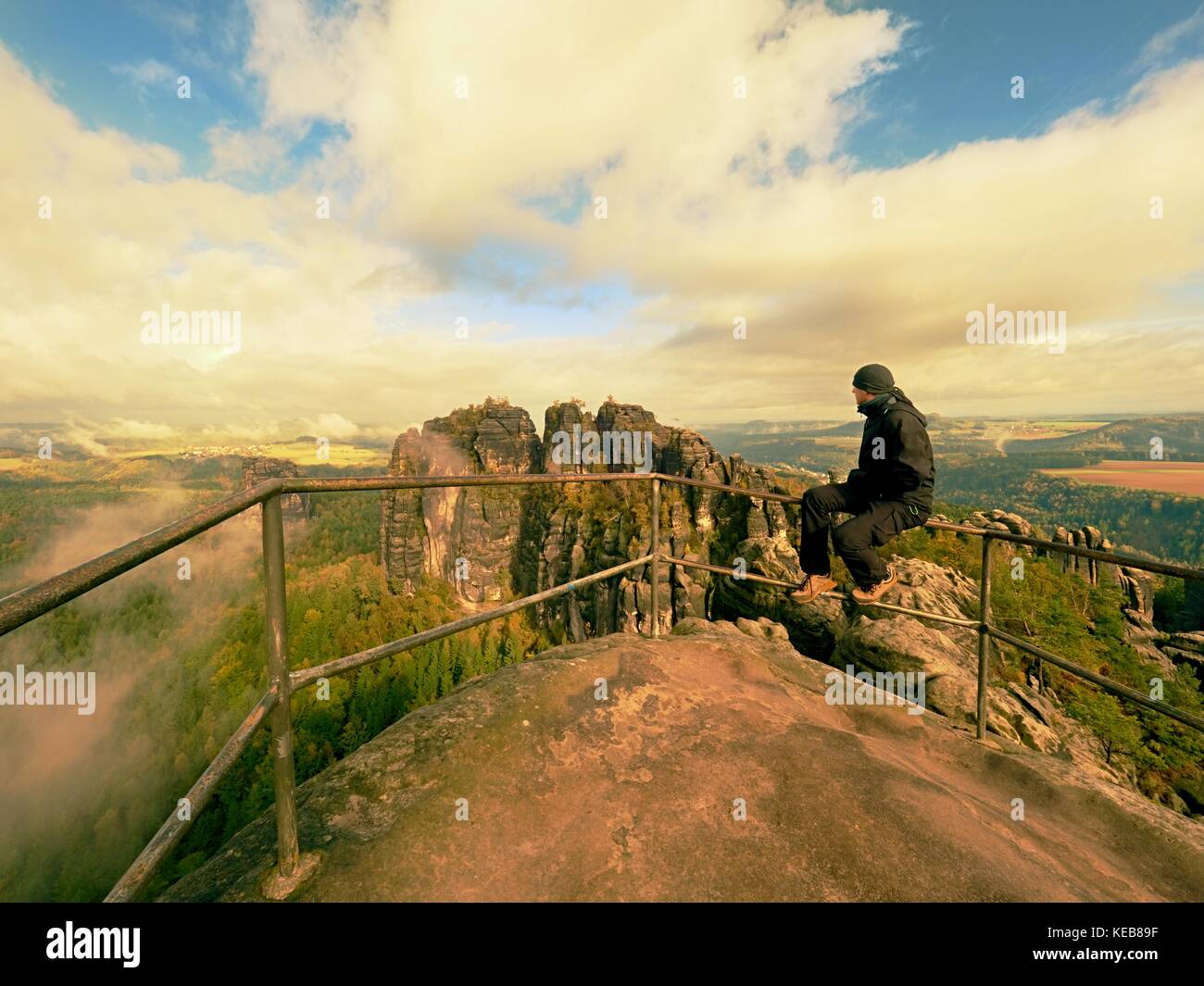 Man hiker at handrail on the peak of sandstone rock watching into misty ...