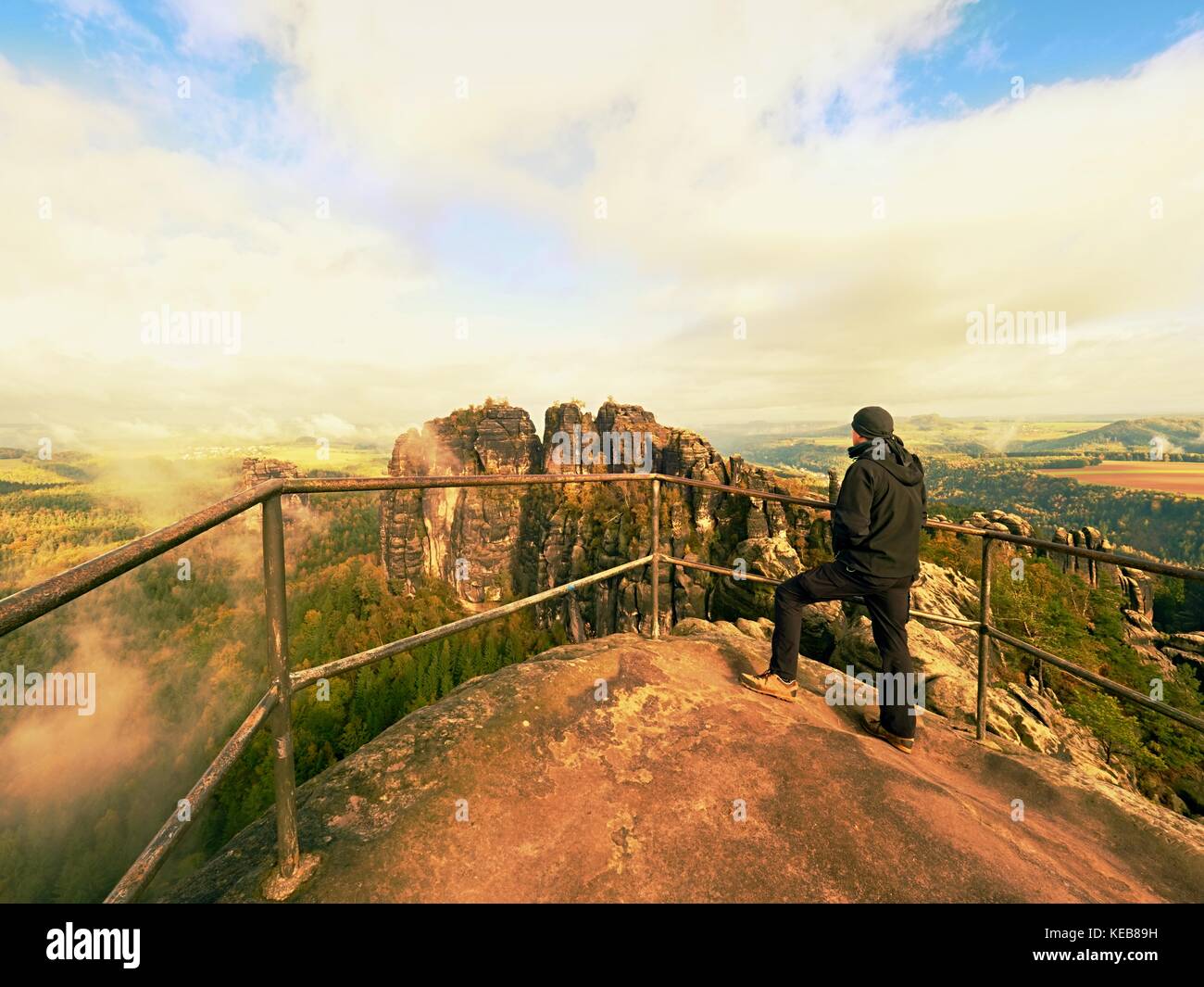 Man hiker at handrail on the peak of sandstone rock watching into misty ...