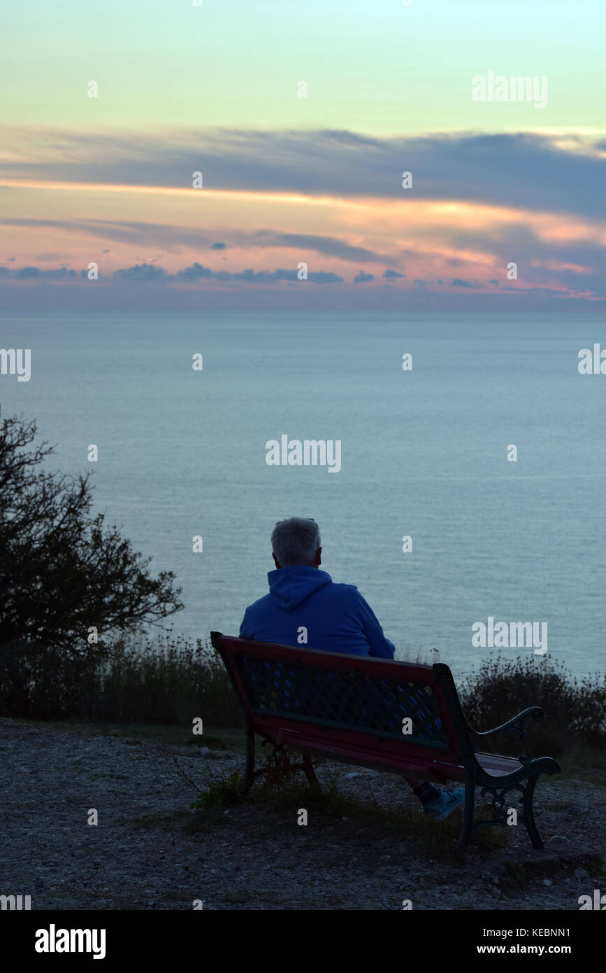 a man sitting on a bench alone at sunset looking out over the sea ...