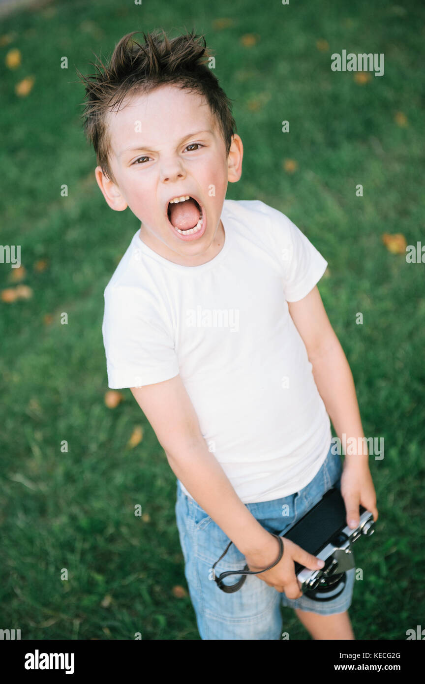 Yelling little boy with film camera on green grass and fallen leaves ...
