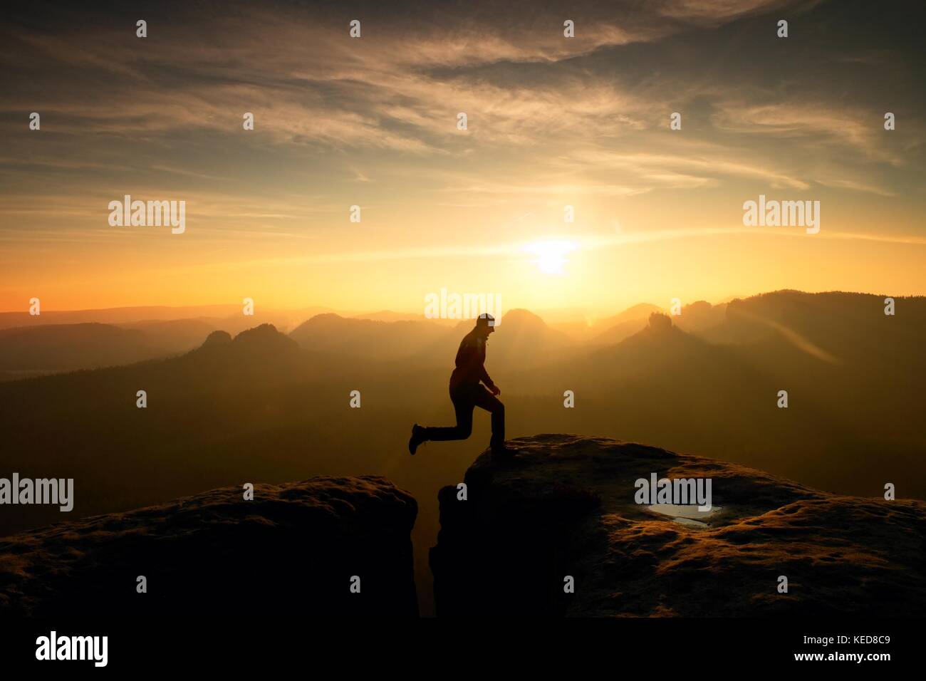 Hiker in black is jumping between mountains peaks. Wonderful daybreak ...