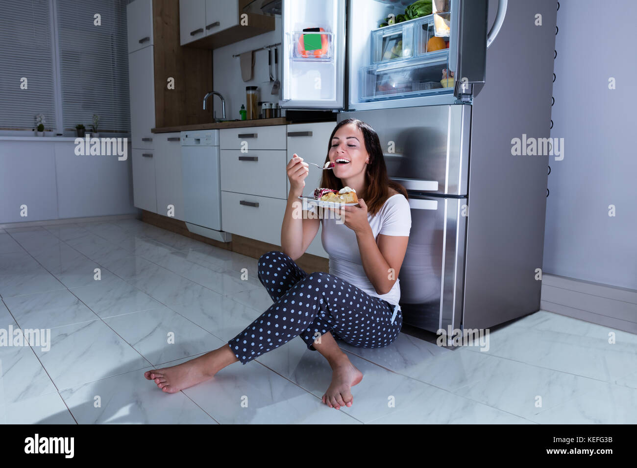 Young Woman Enjoy Eating Sweet Food While Sitting On Floor In Kitchen ...