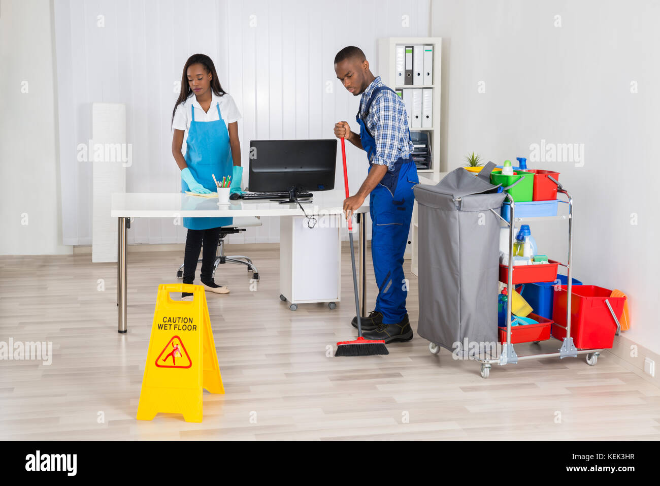 Young Male And Female Cleaners Cleaning Office With Wet Floor Caution ...