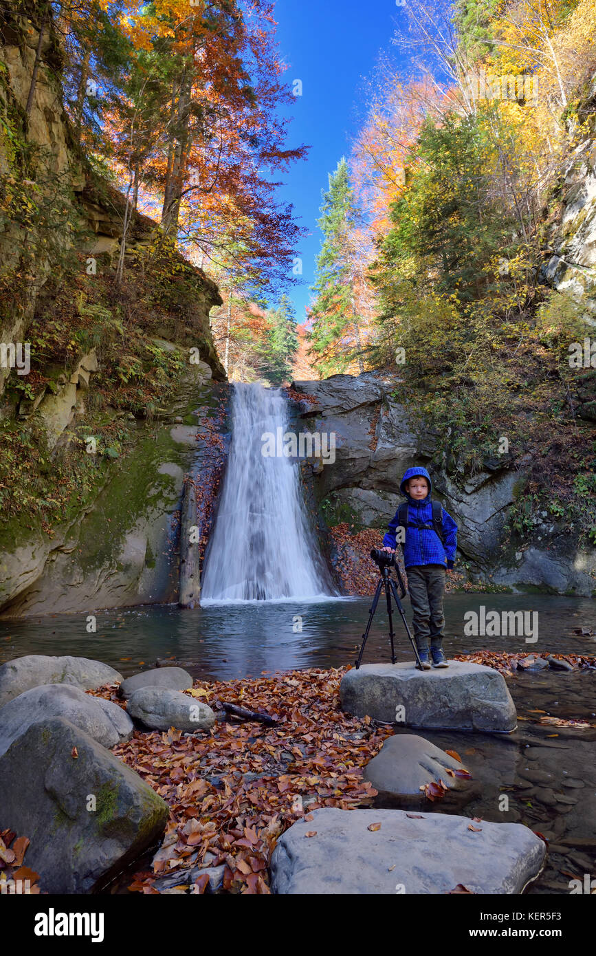 Happy kid near photo camera on tripod with a waterfall in background in ...