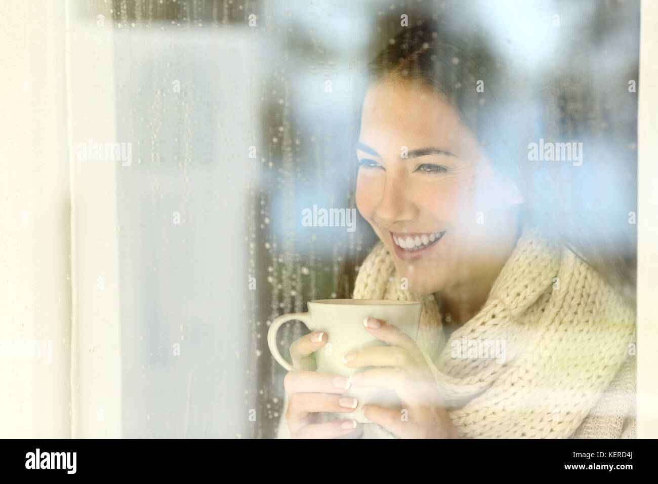 Happy lady looking outdoors through a window holding a coffee cup in ...