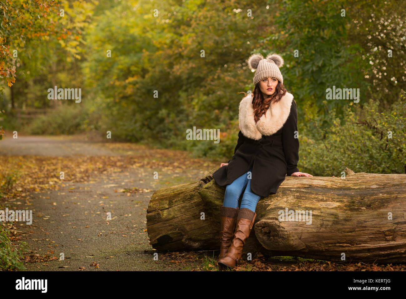 Woman dressed in coat and woollen hat sitting on log in autumn sunshine ...