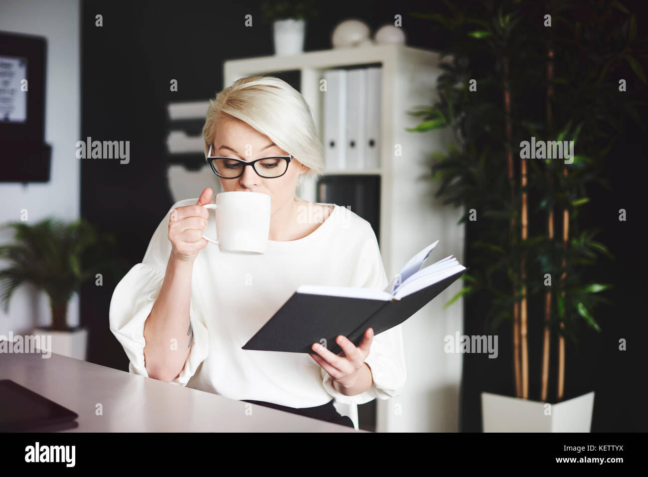 Woman reading book and drinking coffee at her desk Stock Photo - Alamy