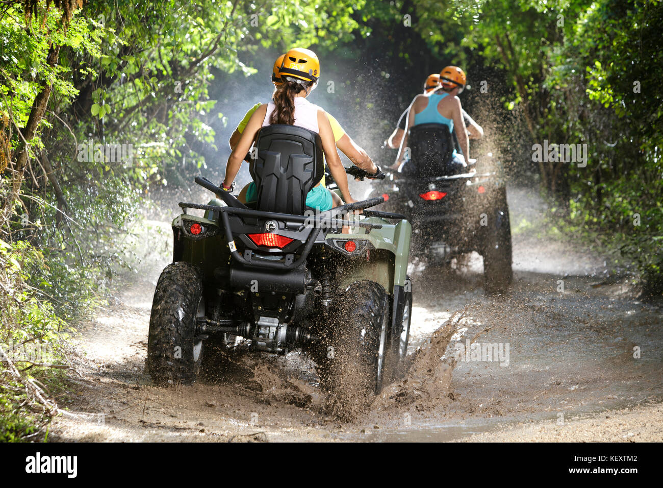 Rear view of couples driving quad bikes through dirt road puddles in ...