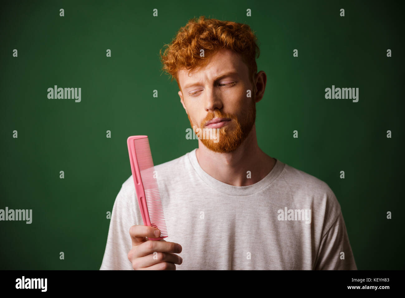 Young readhead beardy man looking at pink comb, over green background ...