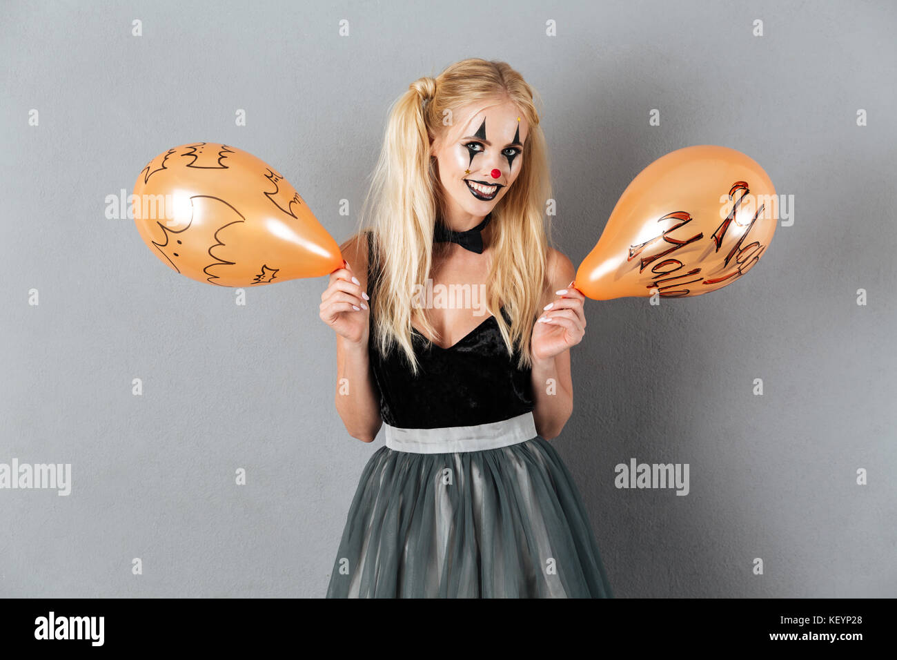 Crazy Blonde Woman In Clown Make Up Holding Halloween Balloon And Smiling Isolated Over Gray
