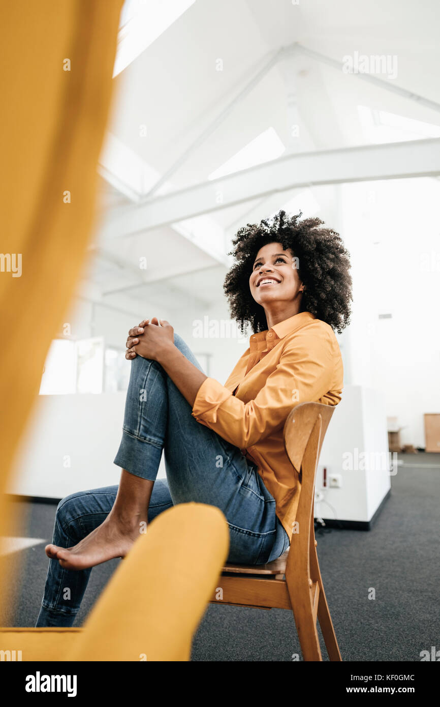 Smiling young woman sitting on chair in office looking up Stock Photo ...