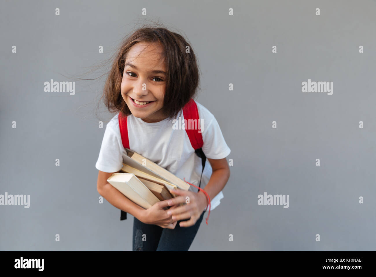 Happy brunette schoolgirl with long hair hugging books and looking at ...