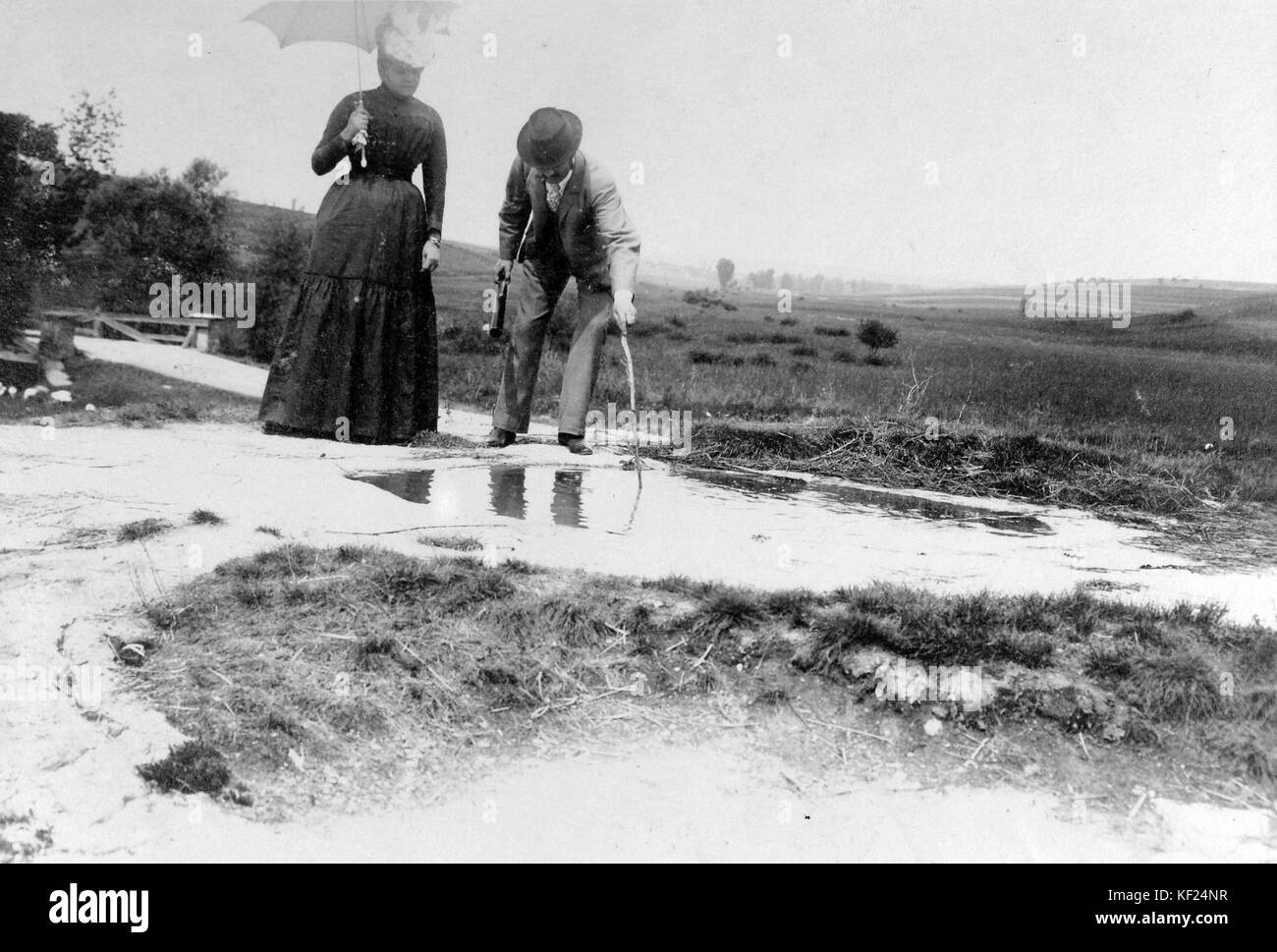This genre painting depicts a woman and man standing near a puddle on a ...