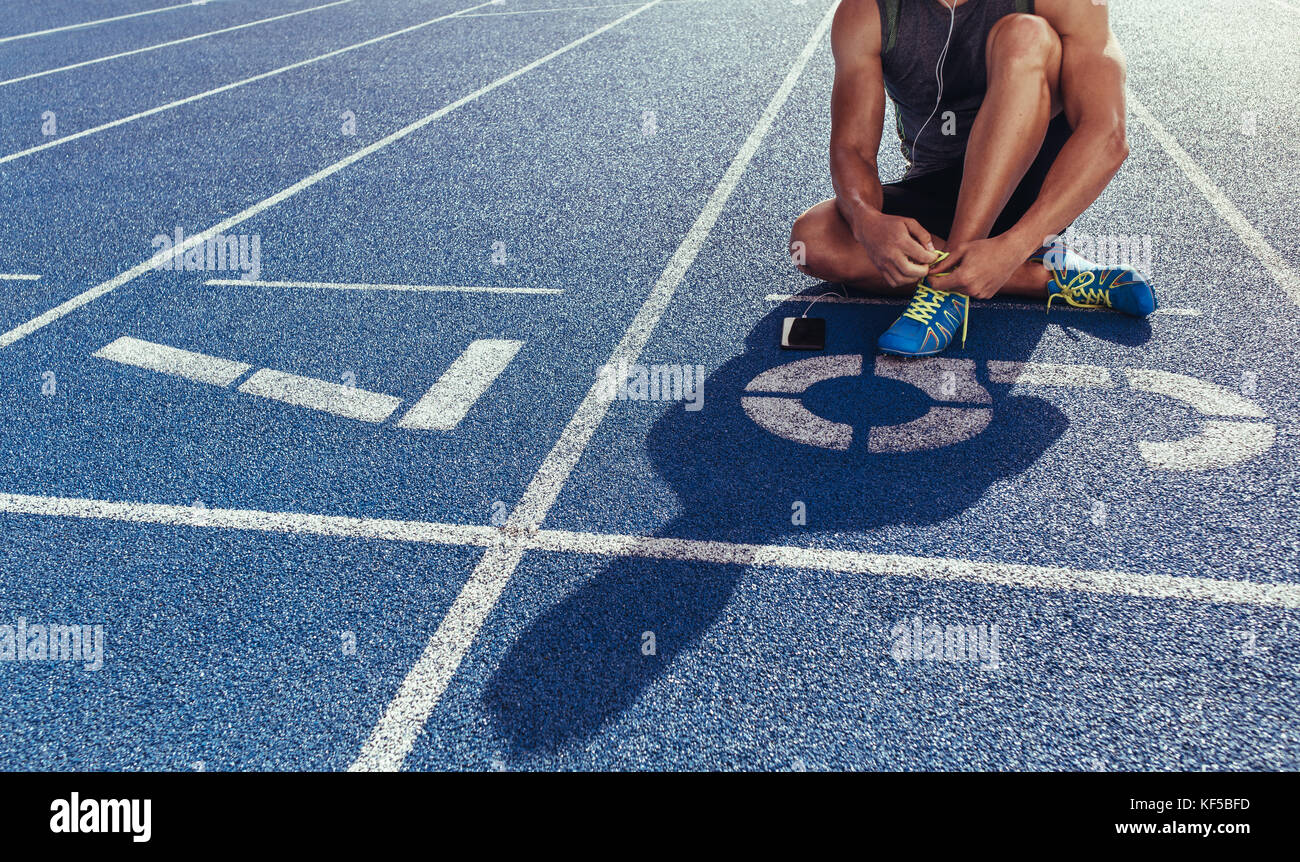 Athlete sitting on a running track tying shoe lace while listening to ...