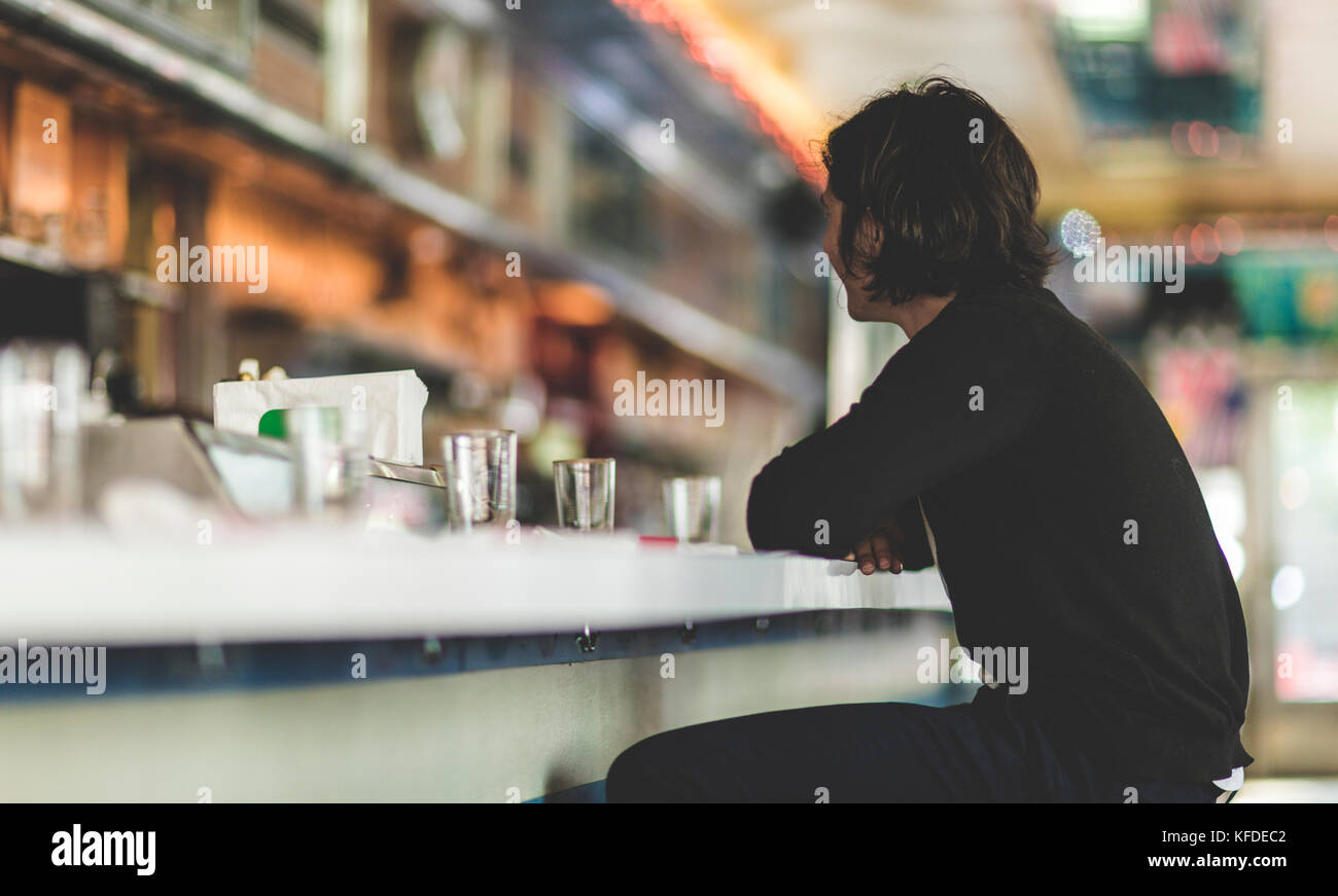 Young man sitting on a stool at a bar counter in a diner, back view ...