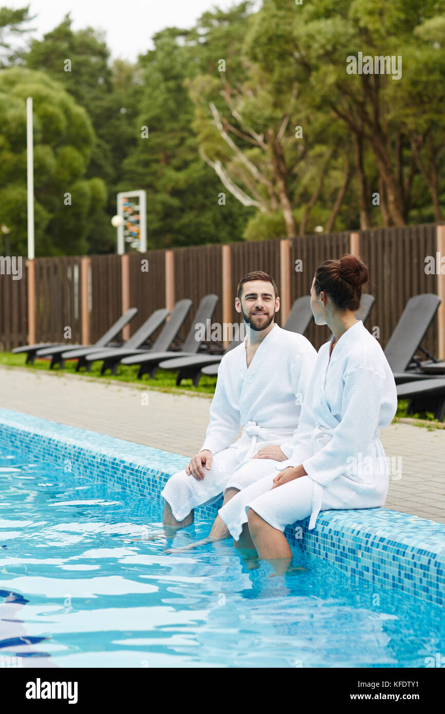 Young couple sitting by swimming-pool with legs in water and talking at ...