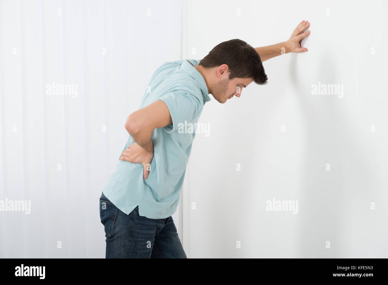 Young Man Having Pain In His Back While Standing Against Wall At Home ...