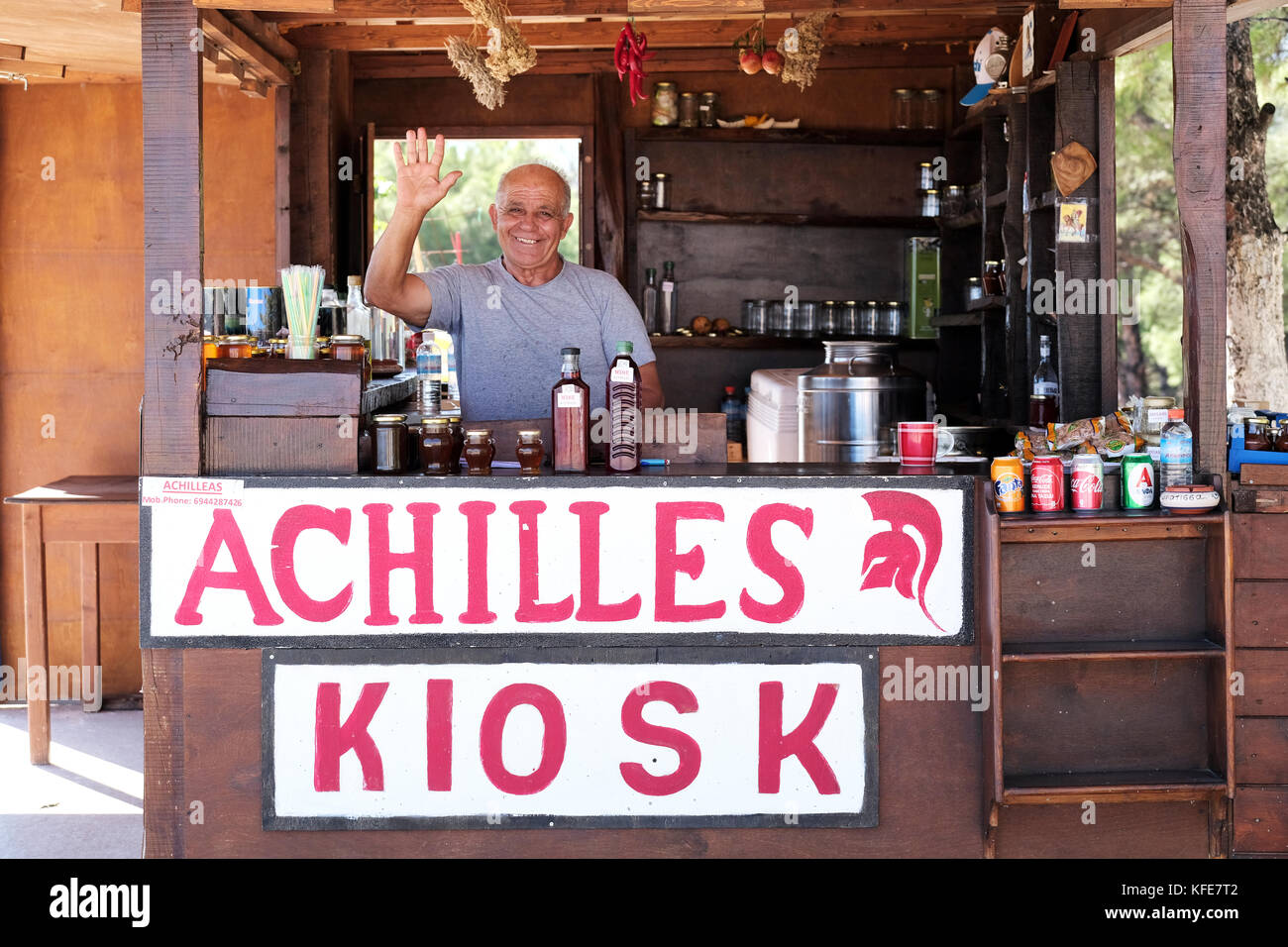 A friendly Greek man waves to the camera from behind the counter of his ...