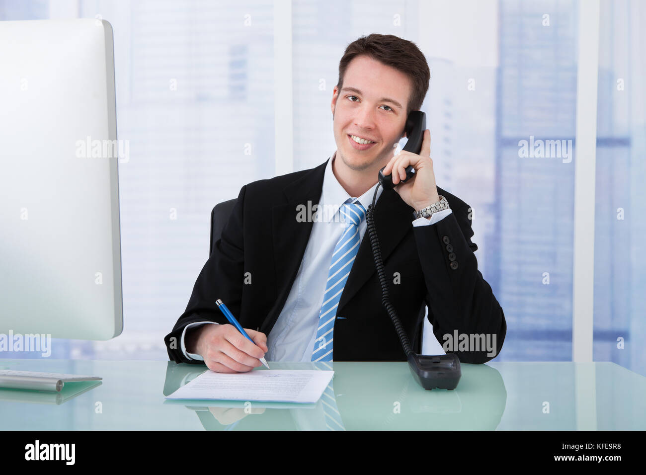 Portrait of young businessman using telephone while writing on document ...