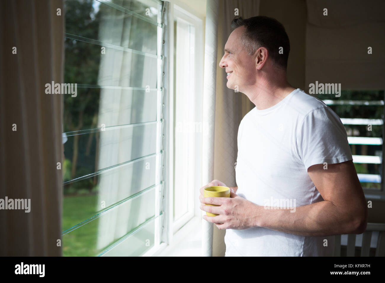 Smiling man looking through window while having cup of coffee in ...