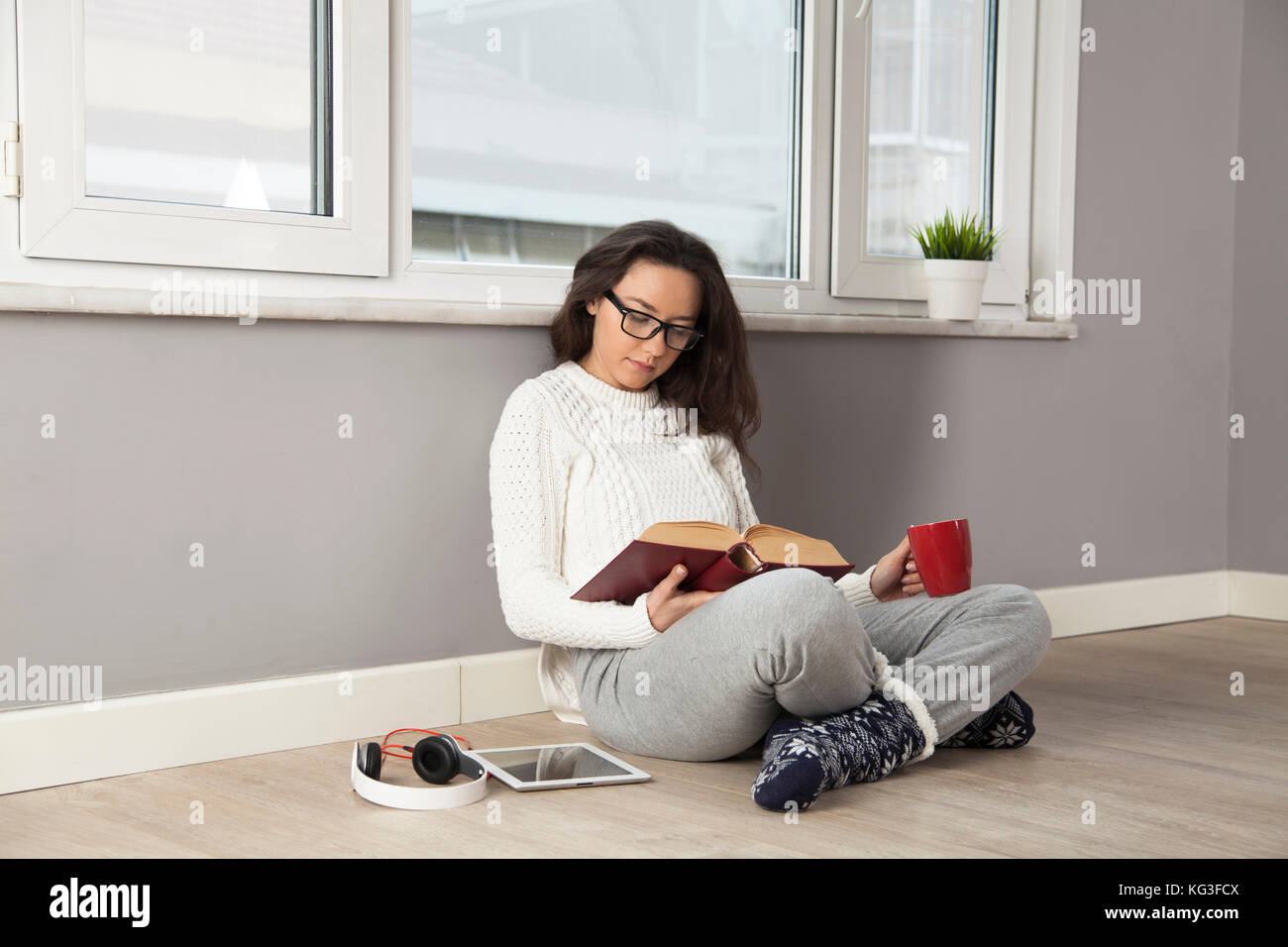 Pensive young woman reading a book while sitting on the floor at home ...
