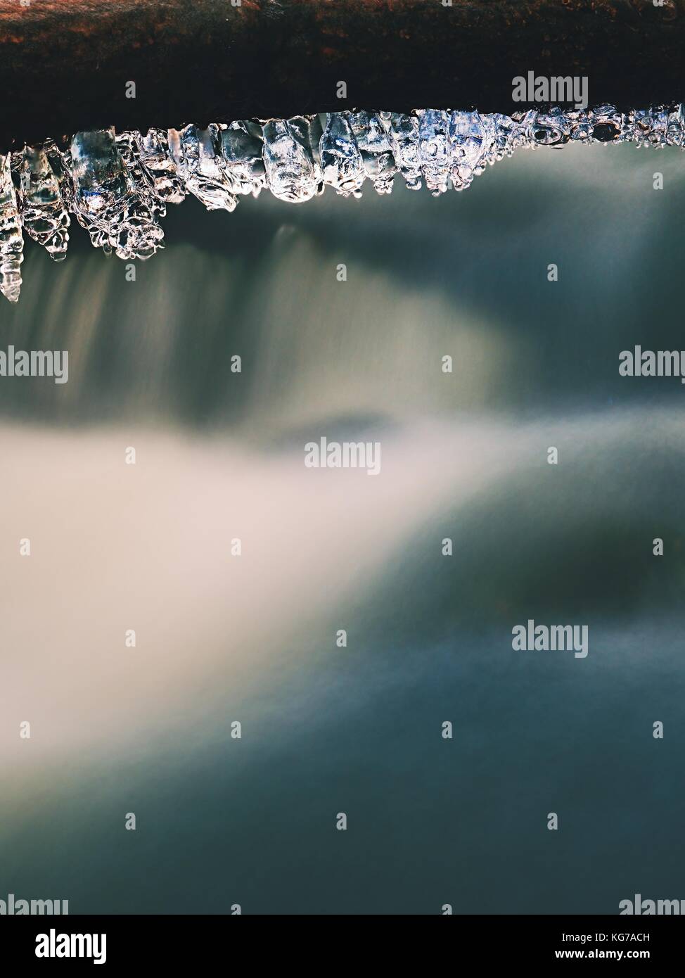 Long icicles hang above dark freeze water of mountain stream. Winter ...