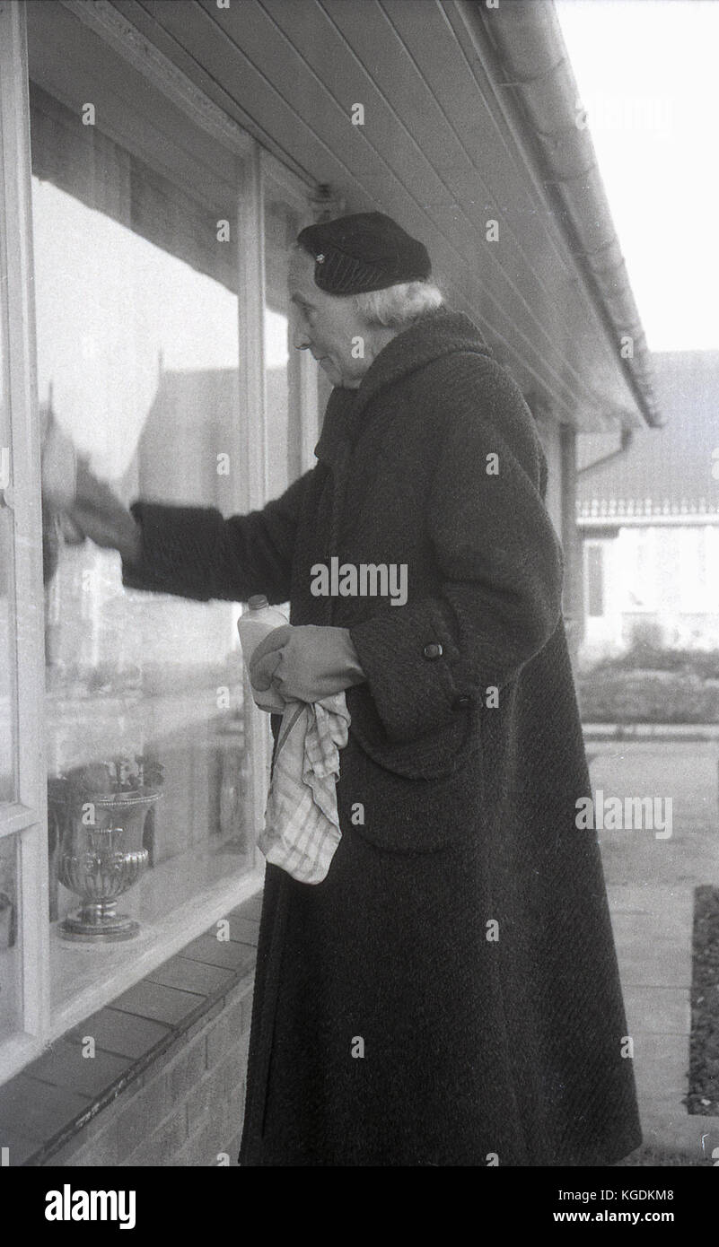 1950s, historical, elderly lady in overcoast outside cleaning a window ...