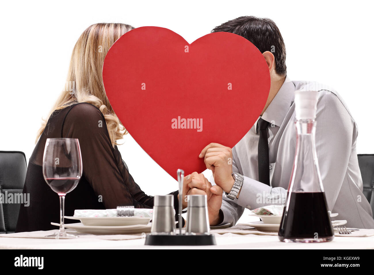 Young couple sitting at a dinner table and kissing behind a red heart ...