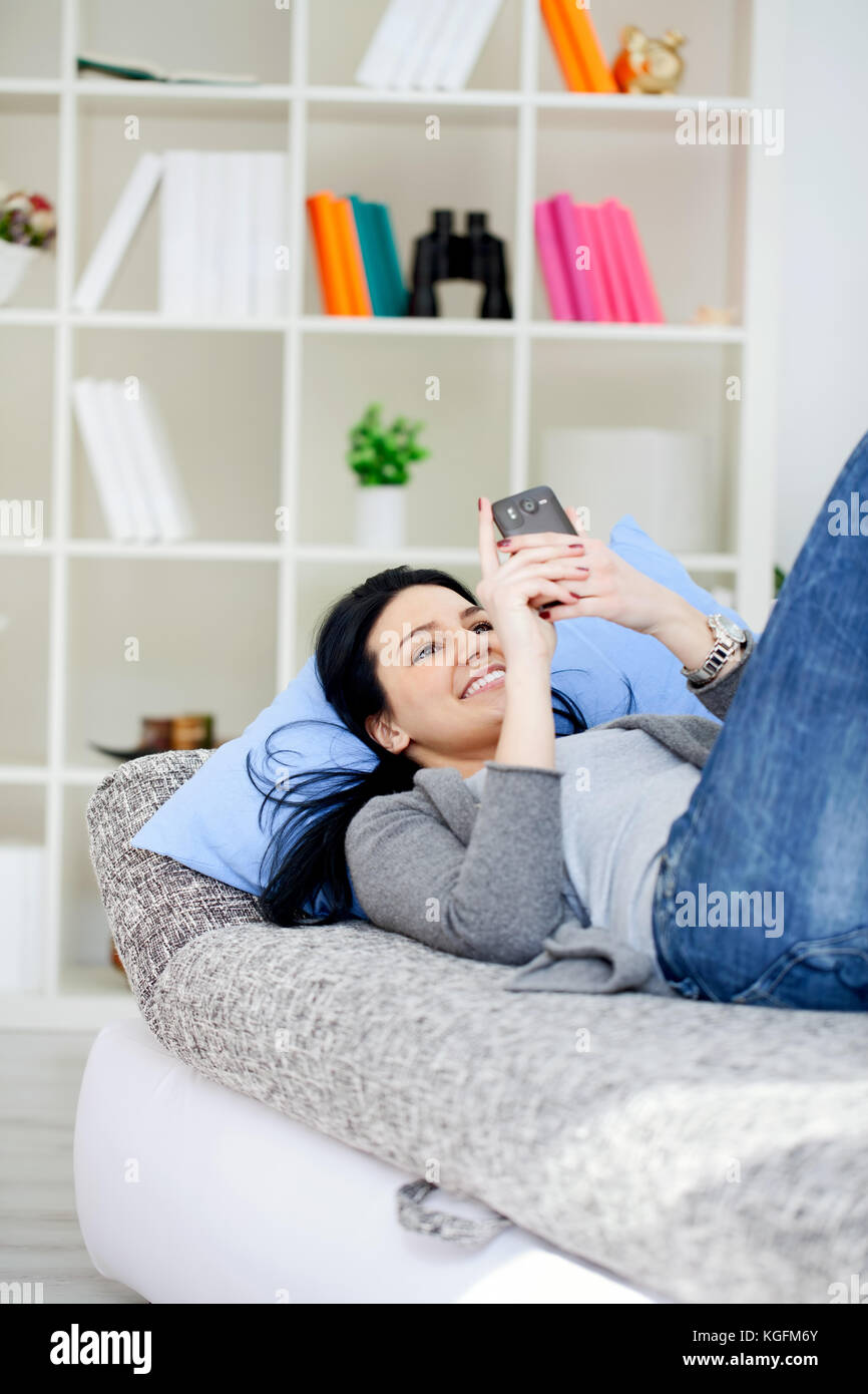 smiling girl lying on sofa and texting message, brightness living room ...
