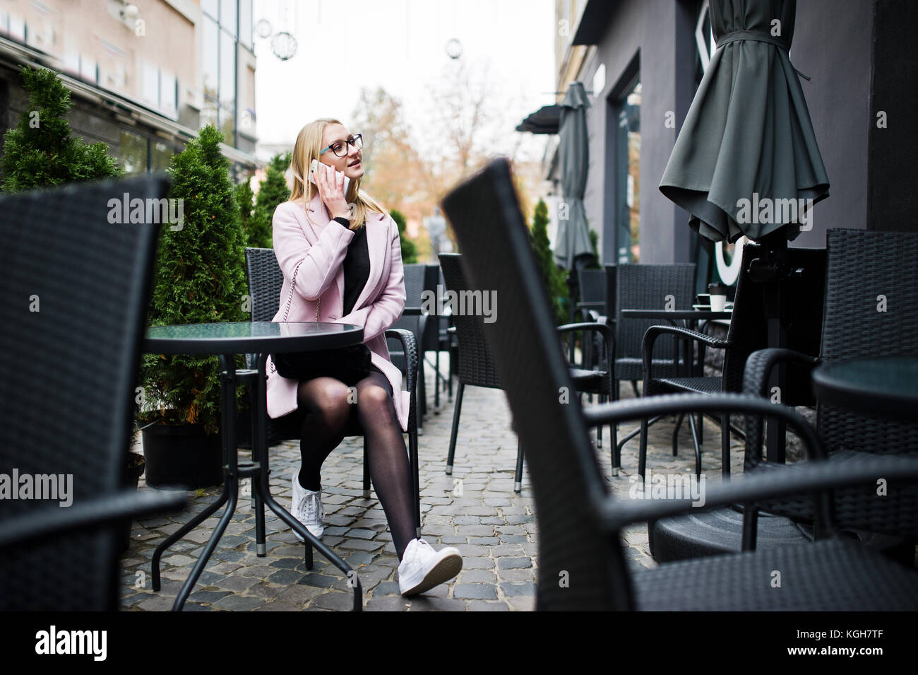 Blonde girl at glasses and pink coat, black tunic sitting at table ...