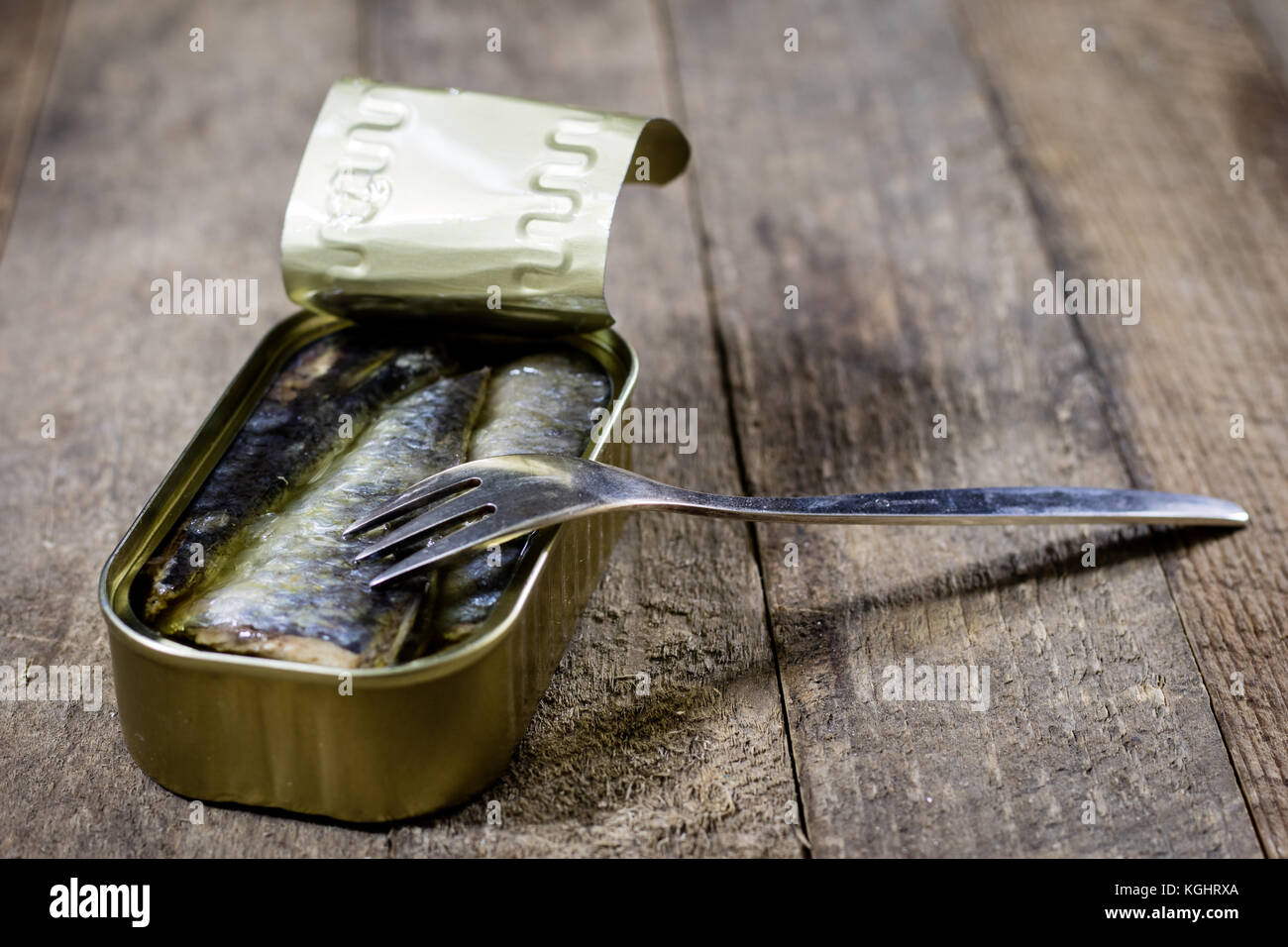 A tin can of fish on a wooden kitchen table. Next to the can fork ...