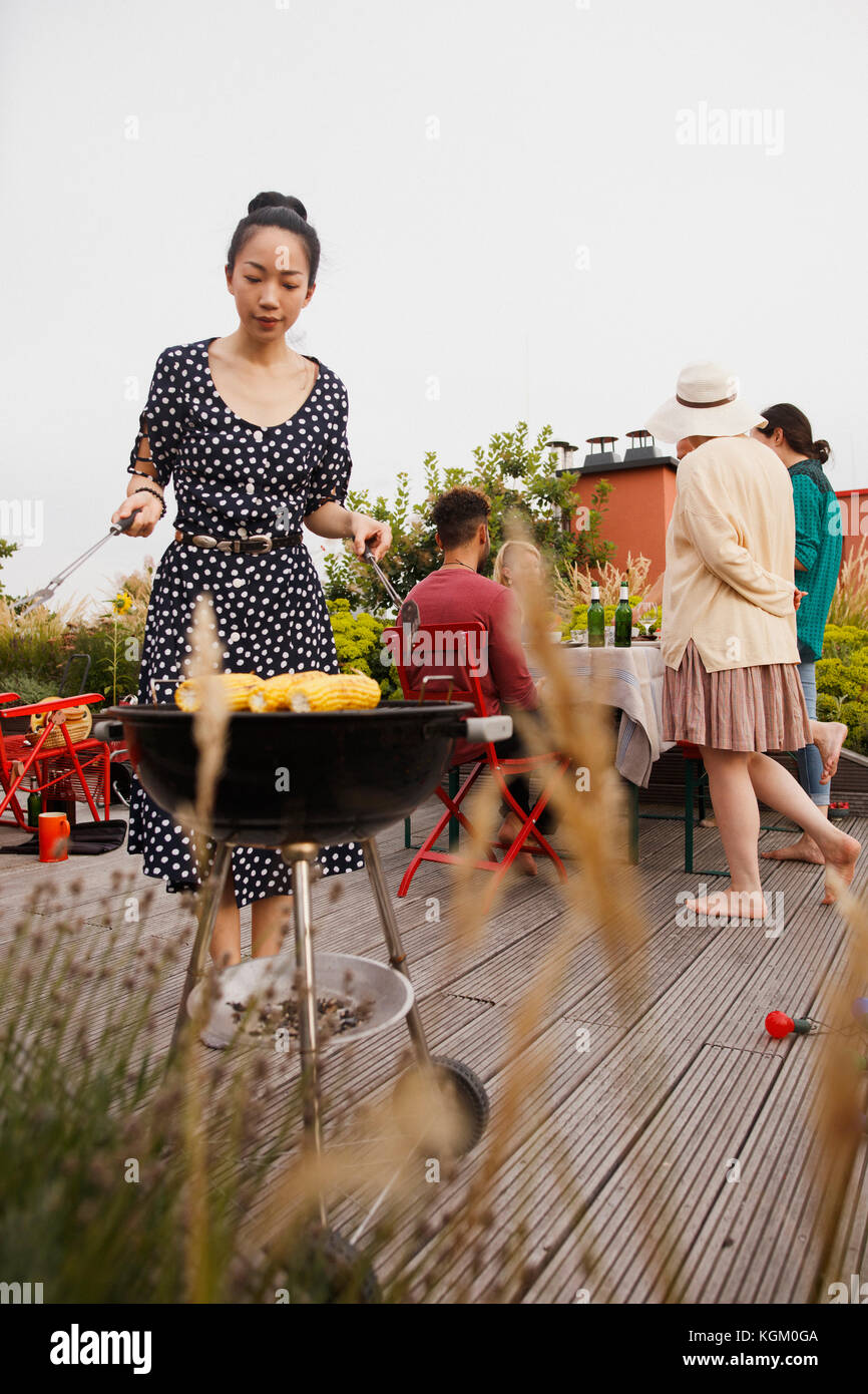 Young woman barbecuing corns while standing on patio with friends in ...