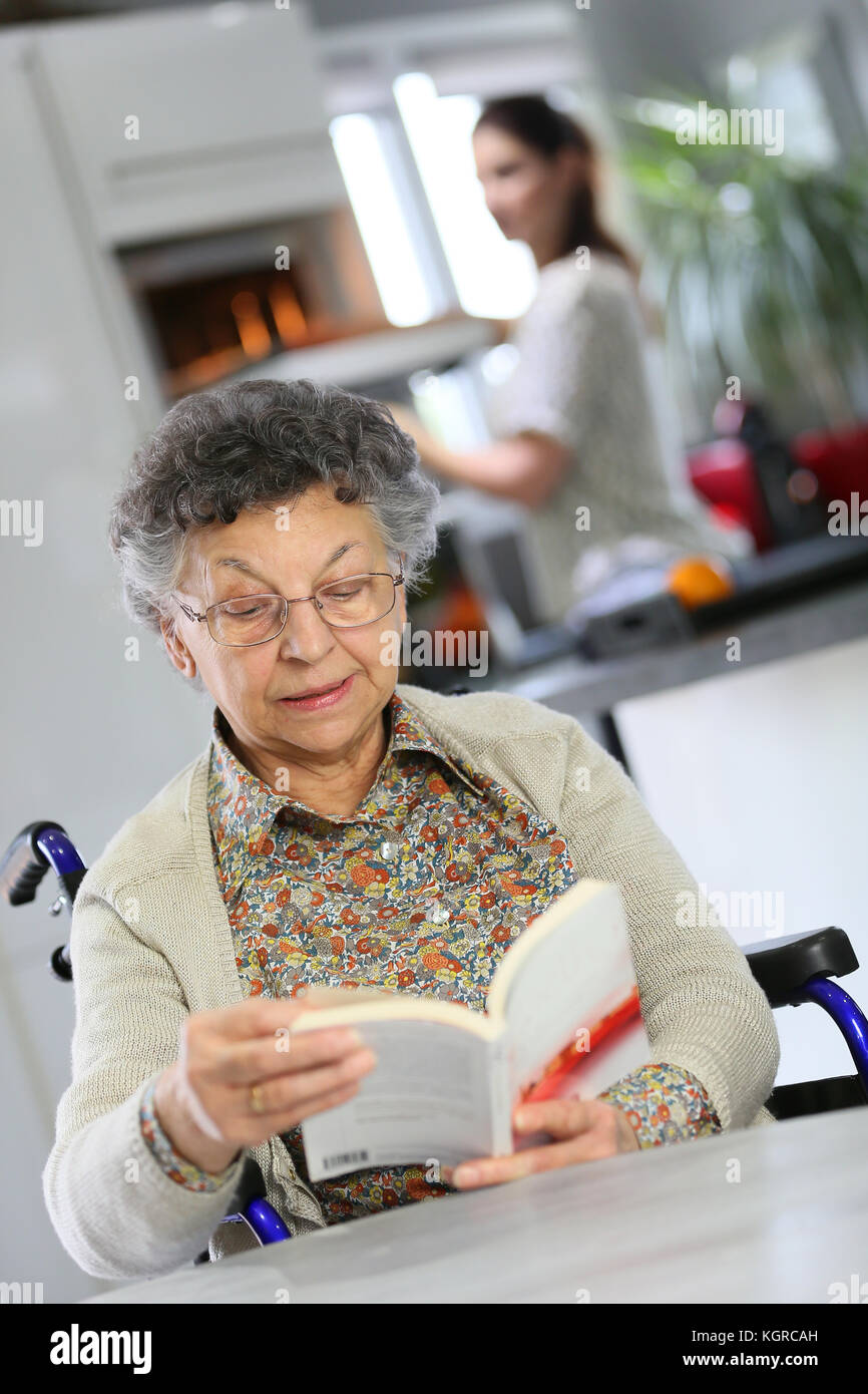 Elderly woman reading book, home helper in background Stock Photo - Alamy
