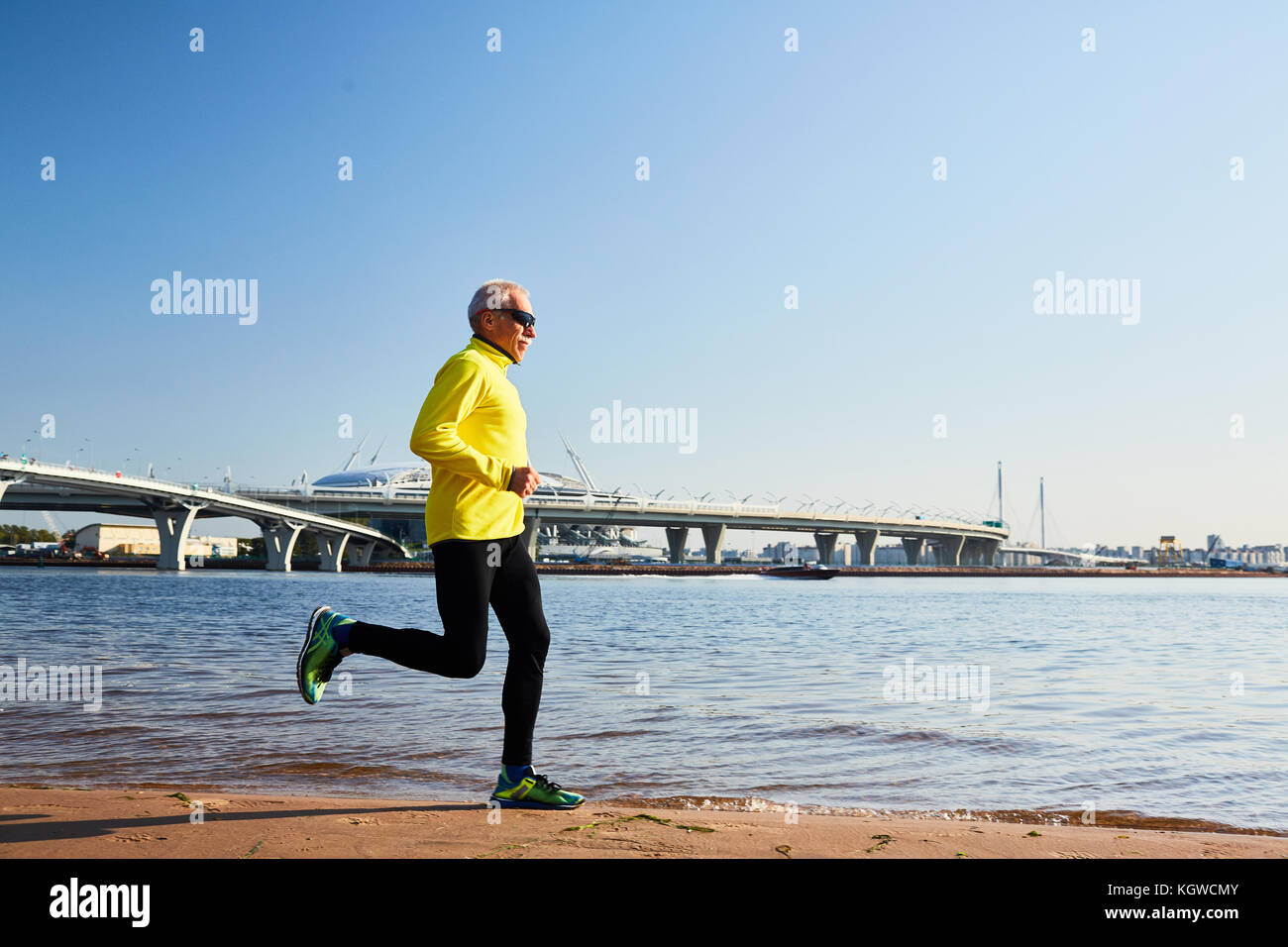 Mature runner working out along river bank with cityscape on background ...
