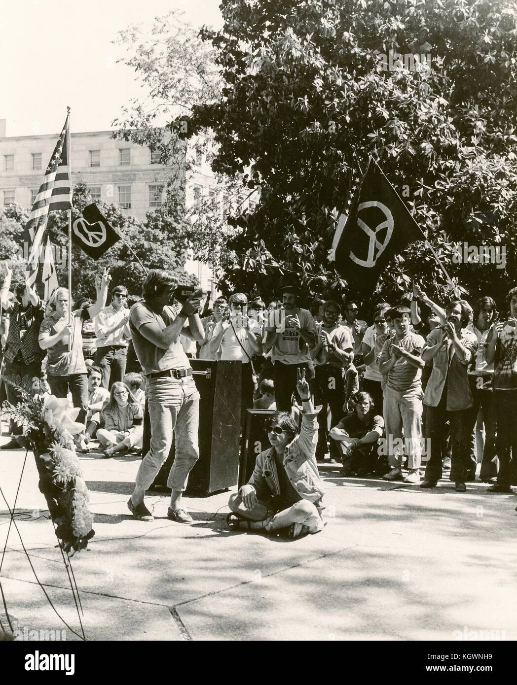 A student wearing hippie attire sits in the middle of a quad and makes ...