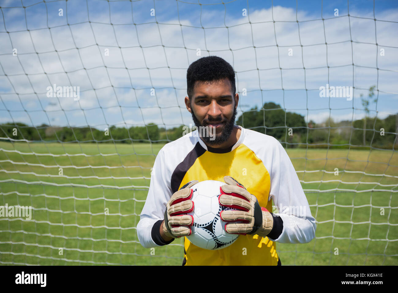 Portrait of smiling goalkeeper with ball standing in front of goal post ...