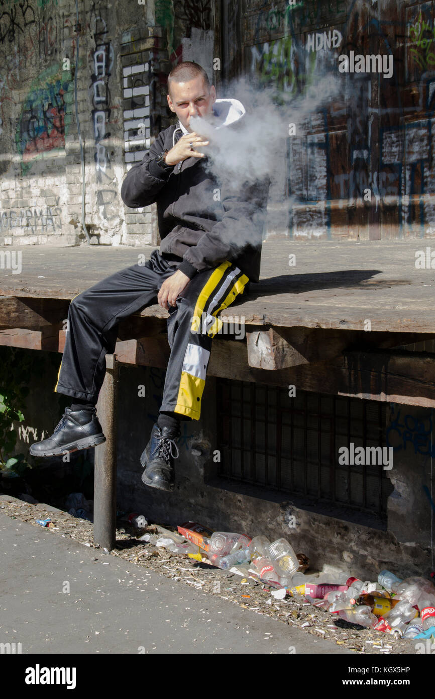 A young man, gopnik, sitting and smoking next to an abandoned warehouse ...