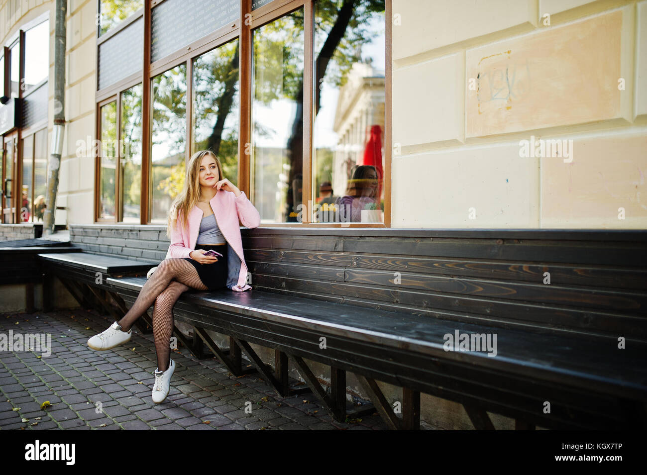 Young blonde girl in black skirt and pink coat sitting on bench at city ...