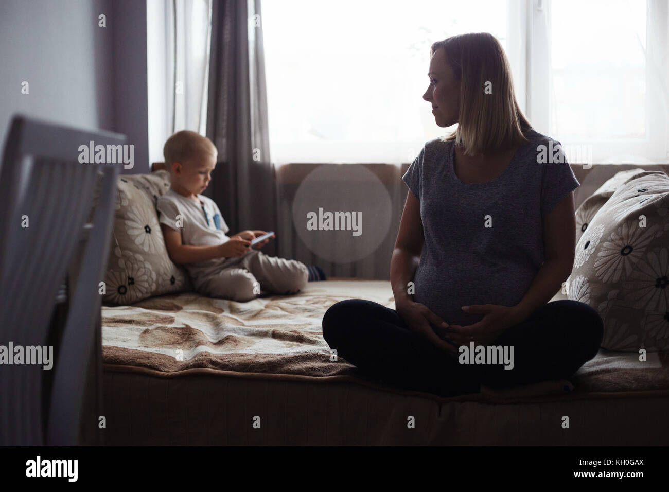 Pregnant Mother Sitting On A Bed Watching Her Son Play A Game On His