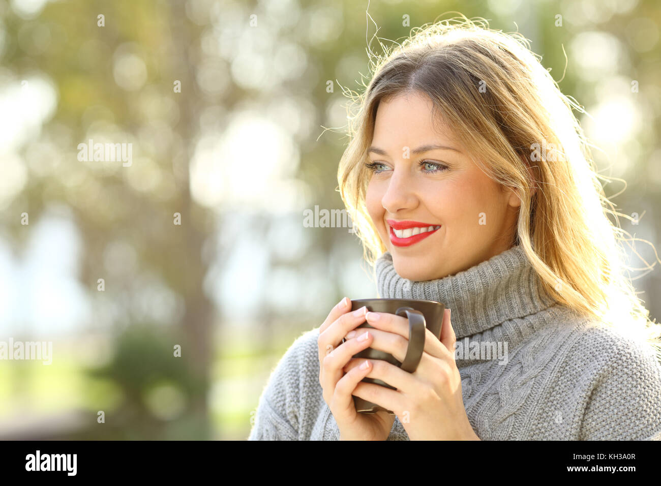 Happy woman wearing sweater thinking holding a cup of coffee in a park ...