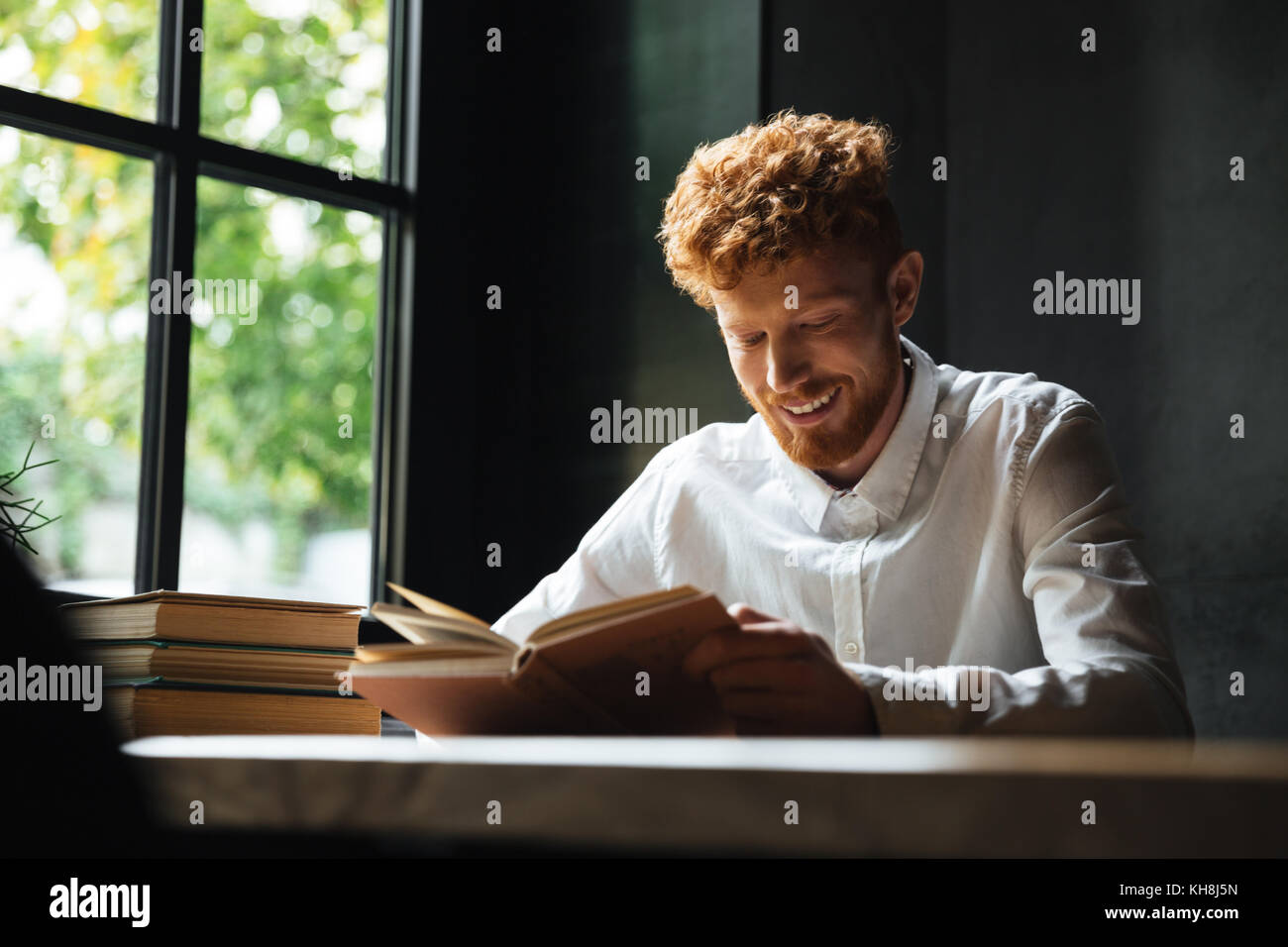Photo of young smiling readhead bearded man in white shirt reading book ...
