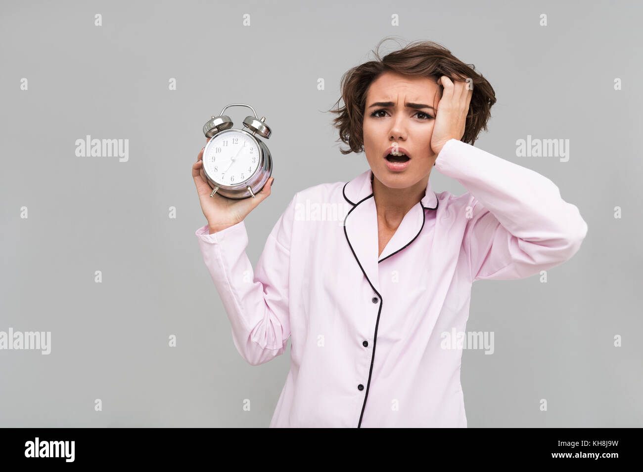 Portrait of a young confused girl in pajamas holding alarm clock and ...