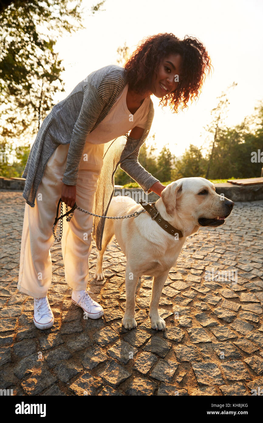 Happy african lady with curly hair hugging her white friendly dog while ...