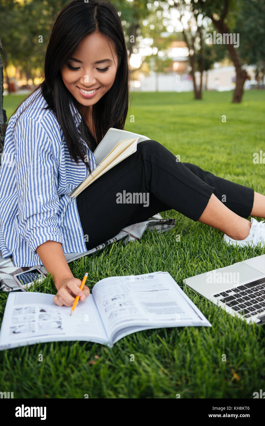 Happy asian female student in striped shirt writing to notebook, while ...